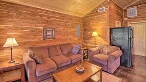 Interior acolhedor de uma cabana com paredes de madeira, um sofá, uma poltrona, uma mesa, um frigorífico e duas lâmpadas.