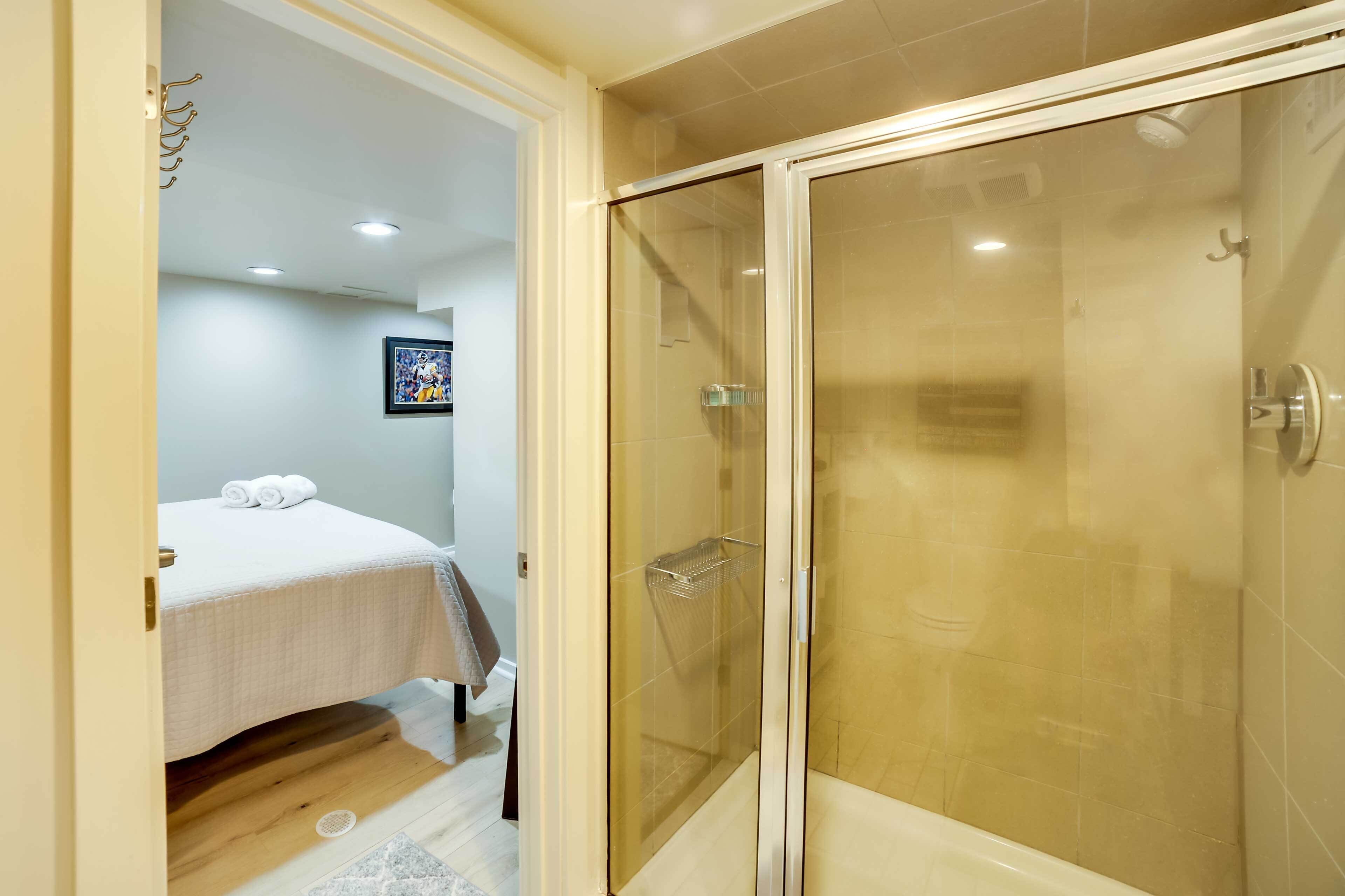 Quarto de hotel com chuveiro em cabine de vidro, cama com colcha branca e quadro na parede.