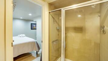 Quarto de hotel com chuveiro em cabine de vidro, cama com colcha branca e quadro na parede.