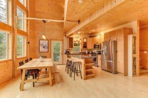 Uma cabana de madeira espaçosa com uma grande mesa de jantar, banquetas, cozinha com eletrodomésticos de aço inoxidável e janelas amplas que oferecem vista para o exterior.