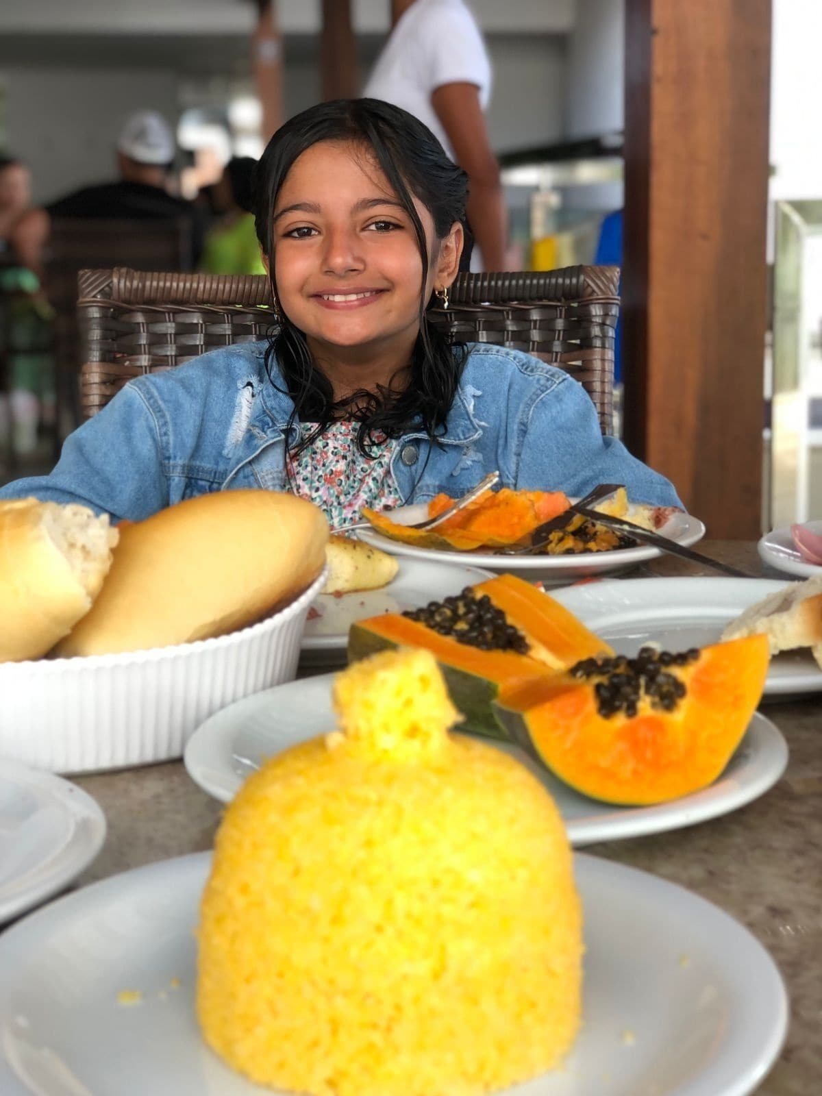 Uma mesa de jantar com diversos pratos, incluindo pão, uma sobremesa amarela e um mamão cortado ao meio.