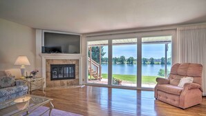 Uma sala de estar ampla com lareira, uma janela grande com vista para um lago, uma poltrona confortável e um sofá.
