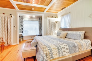 Quarto com cama grande, piso de madeira, um banco e uma janela com cortinas estampadas.
