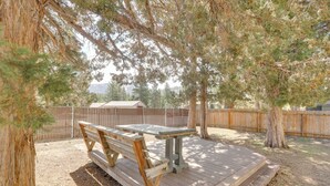 Um deck de madeira com mesa para piquenique e bancos, cercado por árvores e um portão de madeira.