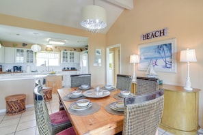 Uma sala de jantar com mesa de madeira posta para quatro, rodeada por cadeiras de vime, um lustre e um letreiro na parede com a palavra 'BEACH'.