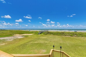 Uma área gramada com um deck de madeira e guarda-corpo de metal, com vista para uma praia com um píer e um veículo azul.