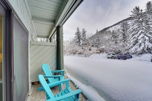 Varanda coberta com cadeiras Adirondack azuis, um caminho de acesso coberto de neve e montanhas ao fundo.