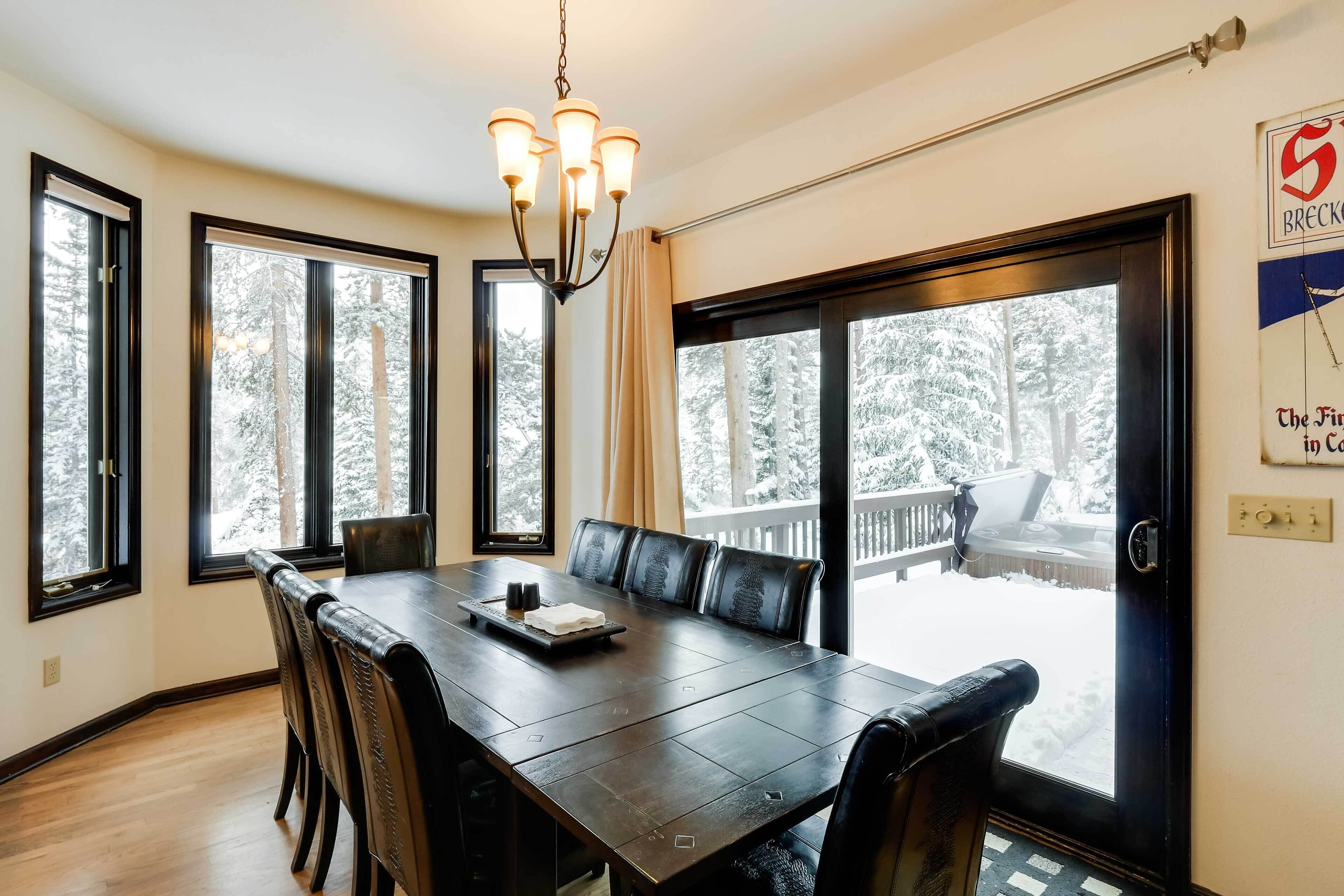 Uma sala de jantar com mesa de madeira escura, cadeiras de couro e um lustre. Grandes janelas oferecem vista para um deck coberto de neve.