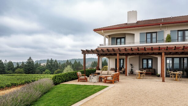 Uma casa de dois andares com varanda coberta, móveis de exterior e um vinhedo ao fundo.