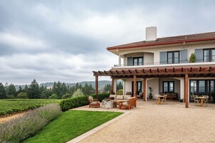 Uma casa de dois andares com varanda coberta, móveis de exterior e um vinhedo ao fundo.