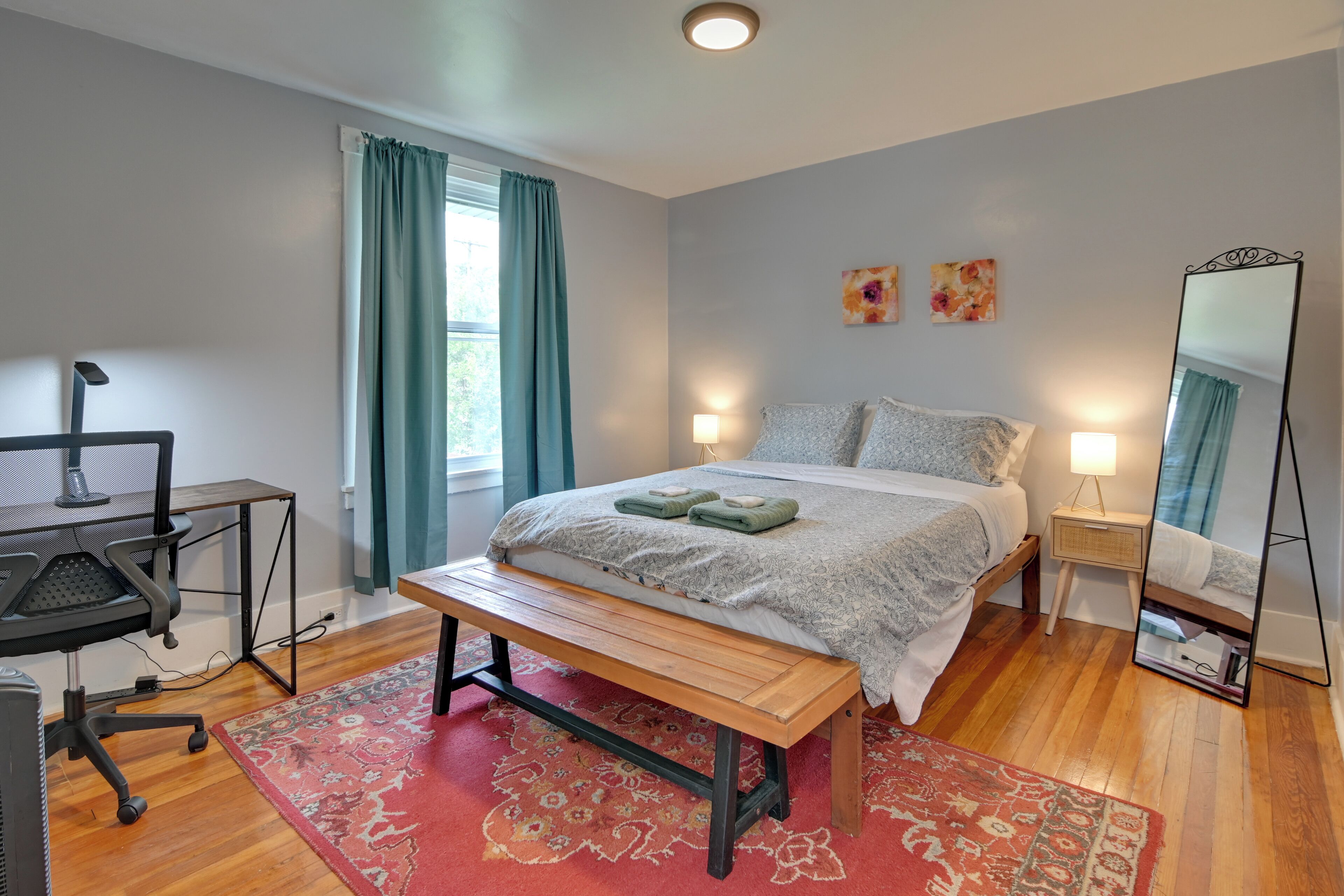 Um quarto com cama, escrivaninha, cadeira, espelho e janela com cortinas.
