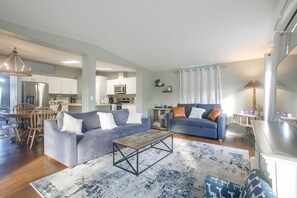 Uma sala de estar moderna com um sofá cinza, um sofá azul, uma mesa de centro de madeira e uma área de jantar com cadeiras e mesa de madeira.