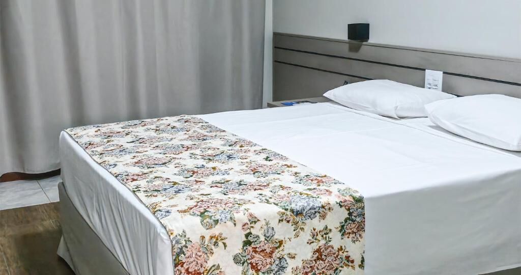 Quarto de hotel com uma cama, cabeceira, luminária fixada na parede e cortina.