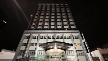 Um edifício alto com design arquitetônico moderno, destacando uma entrada imponente com um toldo de vidro curvo e uma escadaria que leva à entrada.