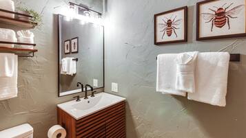 Casa de banho com toucador de madeira, lavatório branco e um espelho grande. Existem duas imagens emolduradas de abelhas na parede, e uma prateleira com toalhas brancas.
