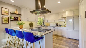 Uma cozinha moderna com armários brancos, uma ilha central com banquetas azuis, eletrodomésticos de aço inoxidável e um exaustor grande.