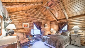 Quarto de cabana de tronco com cama, duas mesas de cabeceira, uma cadeira, uma escrivaninha e um ventilador de teto.
