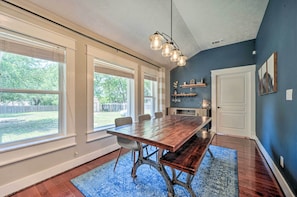 Uma sala de jantar com mesa e cadeiras de madeira, janelas amplas, tapete azul e luminária suspensa.