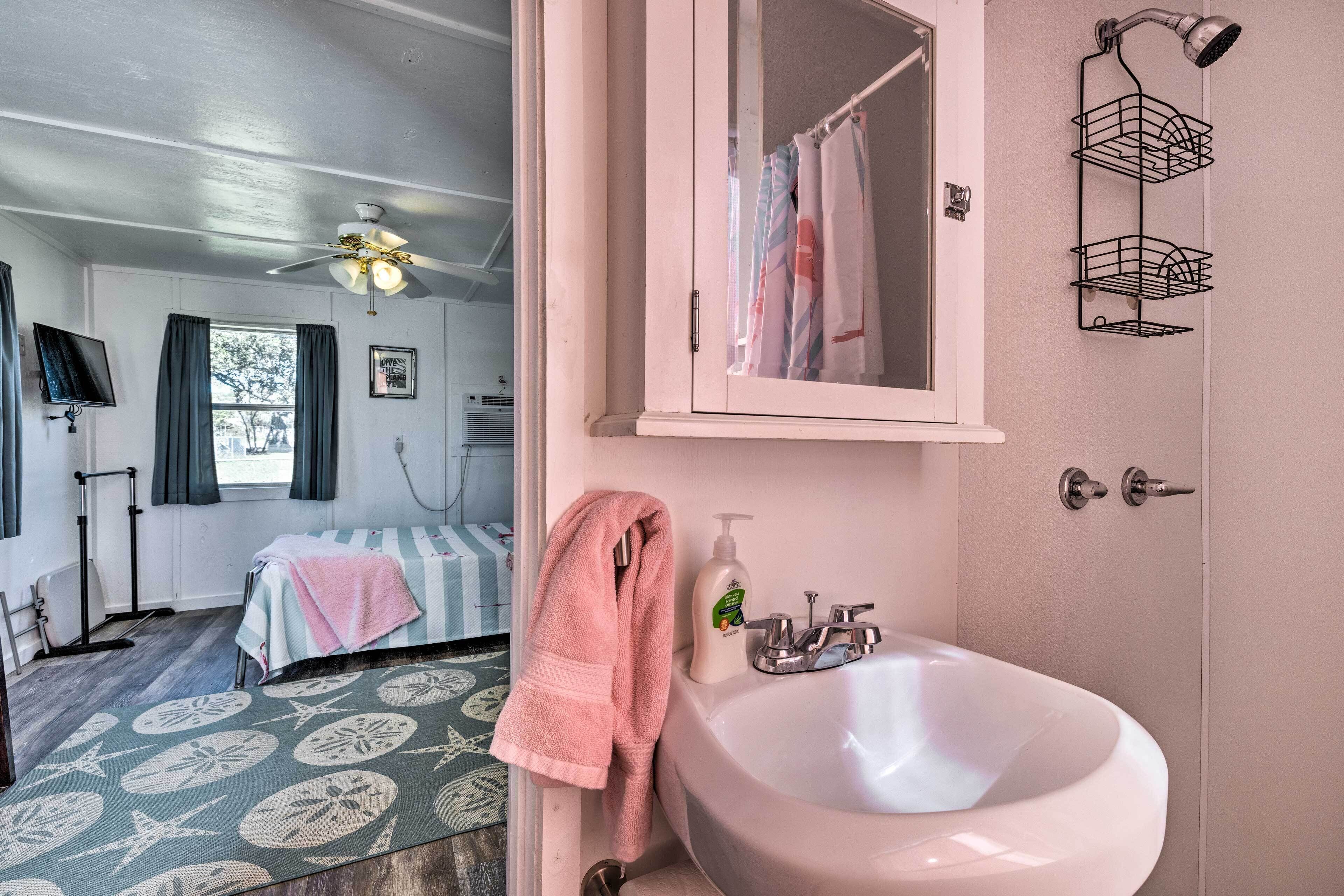 Quarto com cama, ventilador de teto, janela com cortinas e banheiro com pia, espelho e porta-toalhas.
