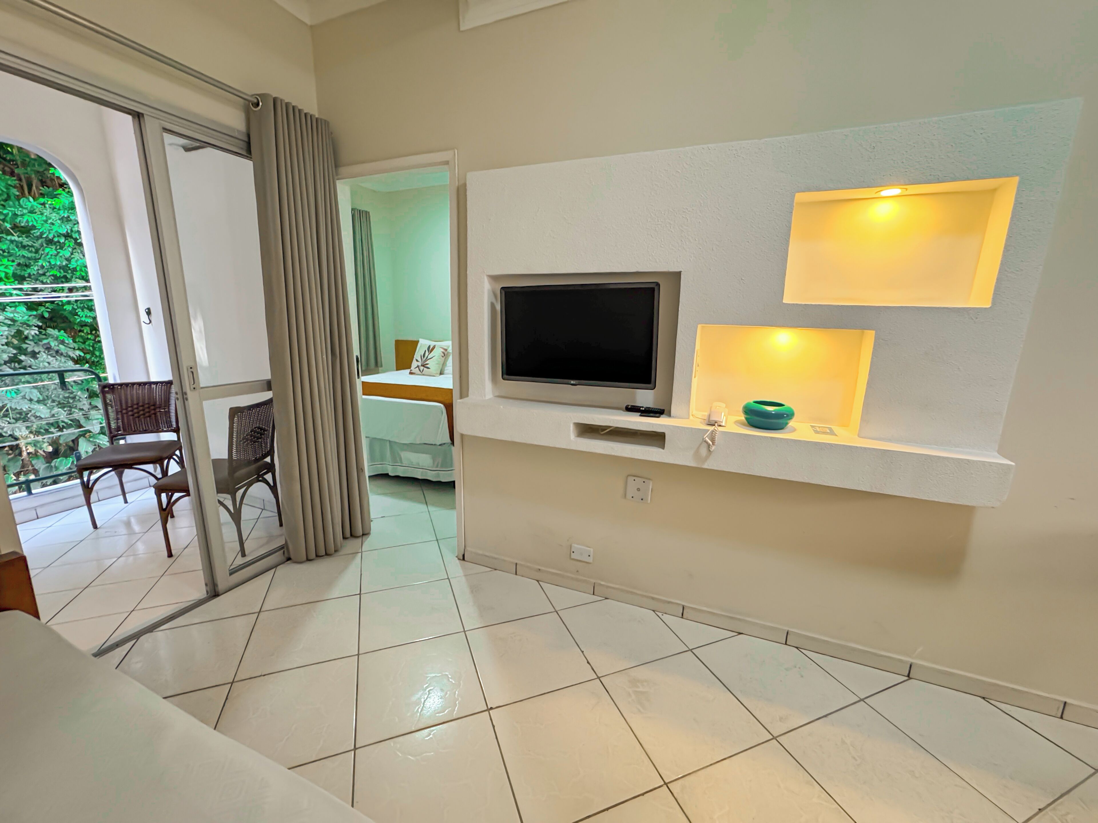 Quarto de hotel com televisão, abajures, uma cama, uma cadeira e uma varanda com vista para o verde.