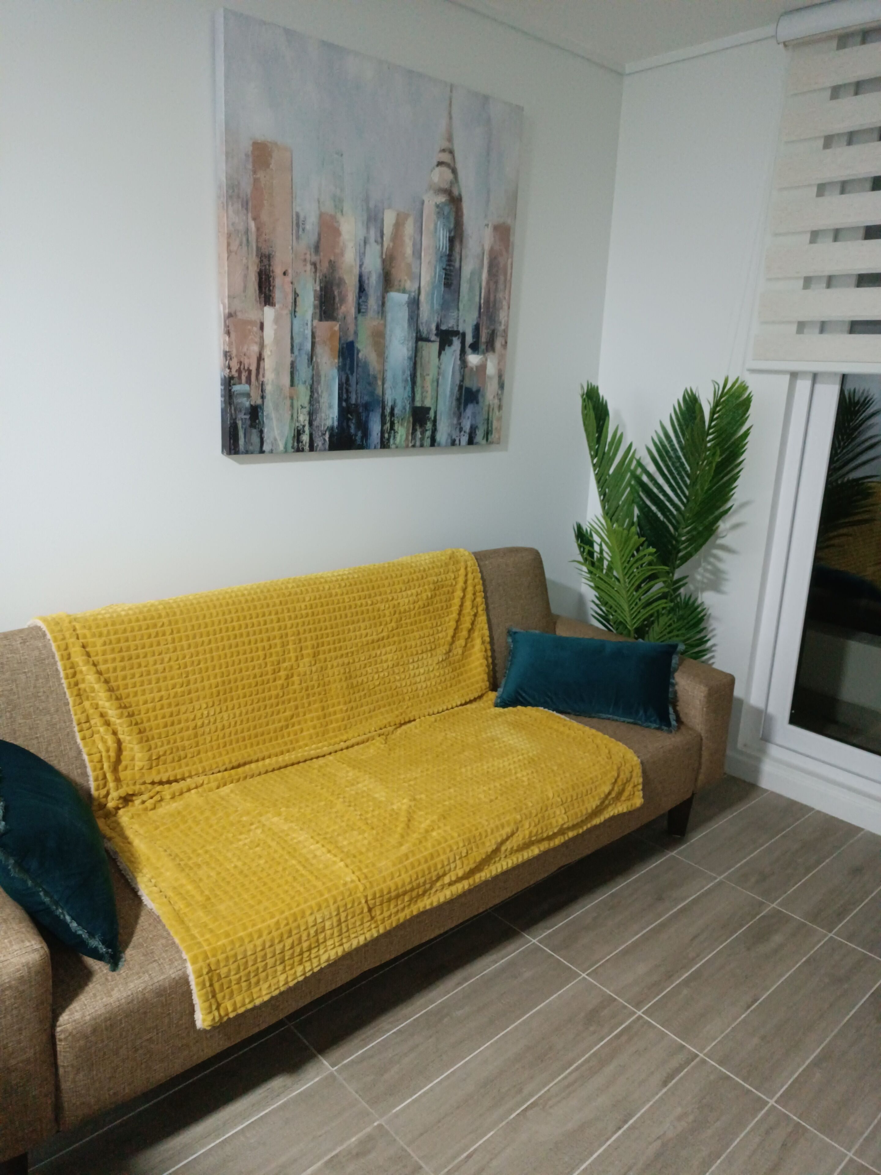 Sala de estar com um sofá coberto por um cobertor amarelo, uma planta verde e uma pintura de paisagem urbana na parede.