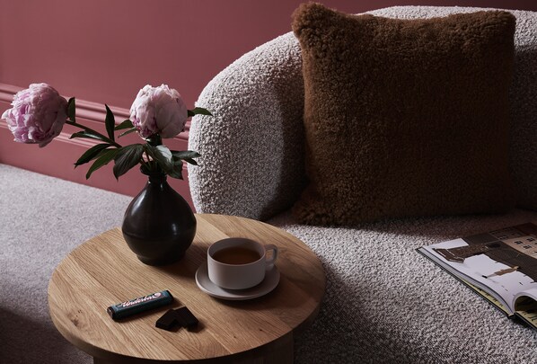 Uma mesa de madeira redonda com uma chávena de café, um vaso escuro com flores cor-de-rosa e uma barra de chocolate.