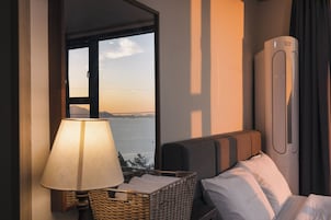 Quarto de hotel com uma cama, uma luminária, uma cesta de vime e vista para o mar.