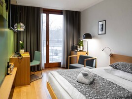 Quarto de hotel com uma cama, uma escrivaninha com telefone, uma cadeira, abajur e uma janela com cortinas.