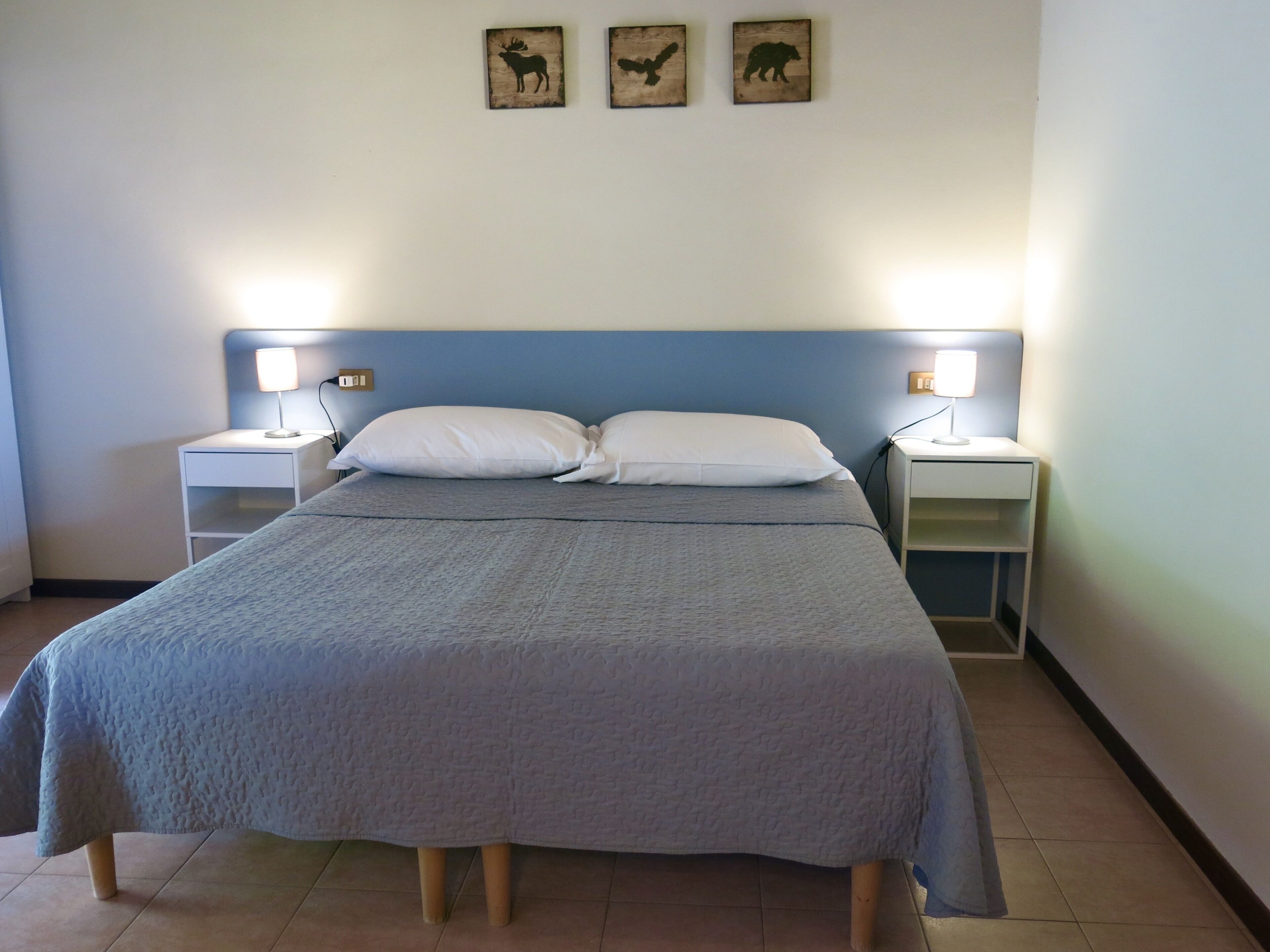 Cama bem arrumada com cabeceira azul e duas luminárias de mesa, um criado-mudo pequeno e três obras de arte com tema de animais na parede.