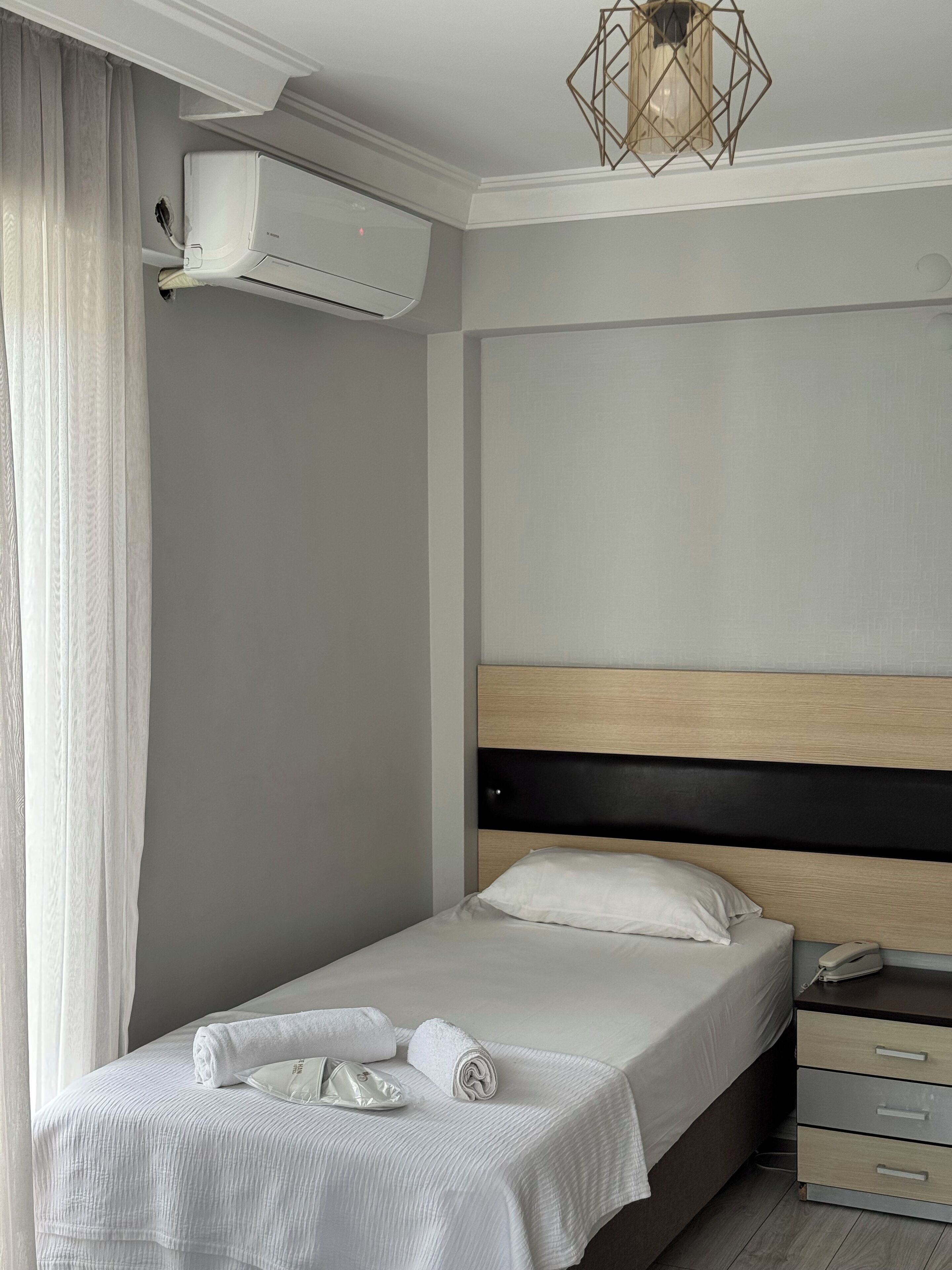 Uma cama bem arrumada com edredom branco e travesseiros, uma mesa de cabeceira com um telefone e um aparelho de ar condicionado fixado na parede.