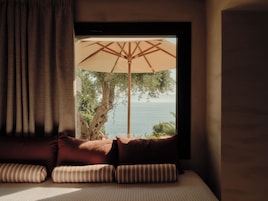 Quarto com uma janela grande, uma cama com almofadas listradas e um guarda-chuva do lado de fora.