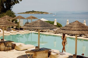 Área de piscina com espreguiçadeiras e guarda-sóis de palha, com vista para um corpo d'água onde se vê um veleiro ao longe.