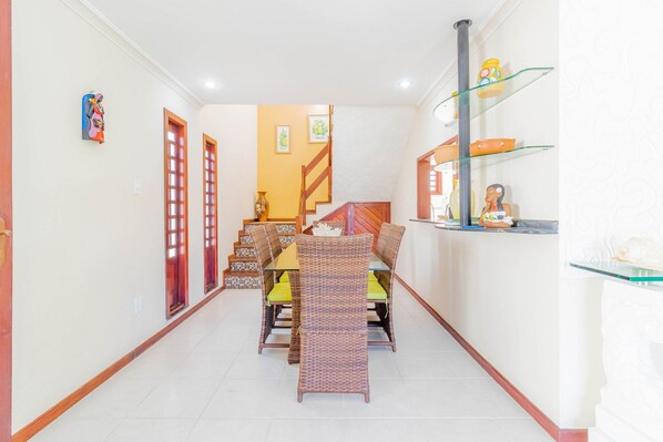 Corredor com área de refeições, cadeiras de vime e uma mesa, escada e um aparador com objetos decorativos.