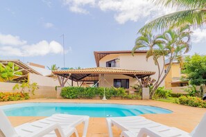 Uma área de piscina com duas cadeiras de praia brancas, um alpendre coberto e uma casa com telhado cerâmico.
