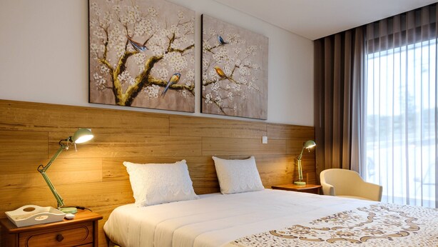 Quarto de hotel com cabeceira de madeira, cama grande com roupa de cama branca, duas pinturas de pássaros penduradas na parede, uma janela com cortinas e uma mesa de cabeceira com abajur.
