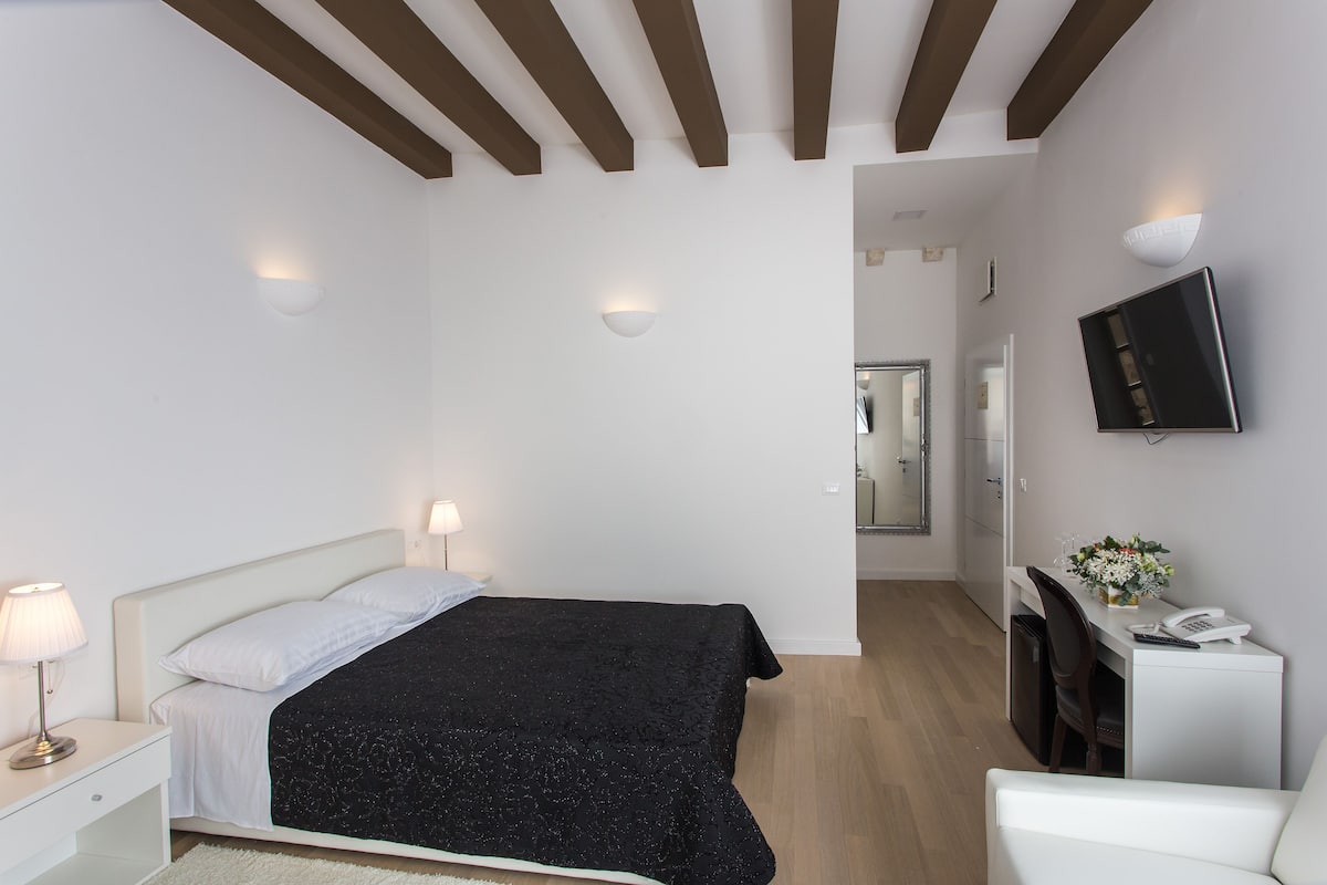 Quarto com cama, luminárias de cabeceira, escrivaninha com cadeira, televisão e pequena mesa com um vaso de flores.