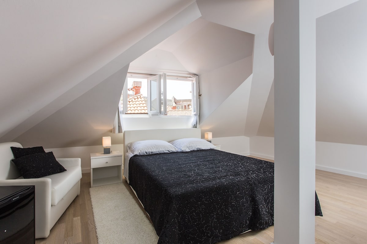 Quarto moderno com teto inclinado, uma cama com colcha preta, mesa de cabeceira branca com abajur e uma cadeira branca.
