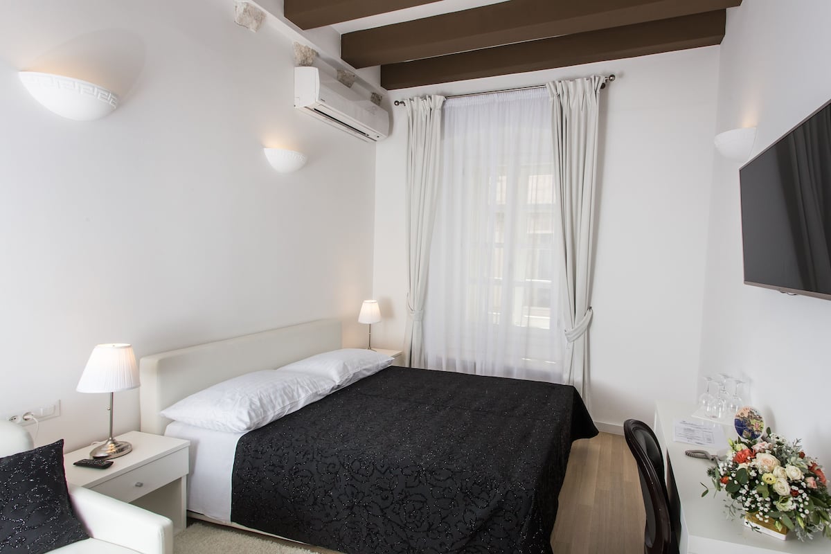 Quarto de hotel com cama, luminárias laterais, escrivaninha com cadeira, televisão e janela com cortinas.