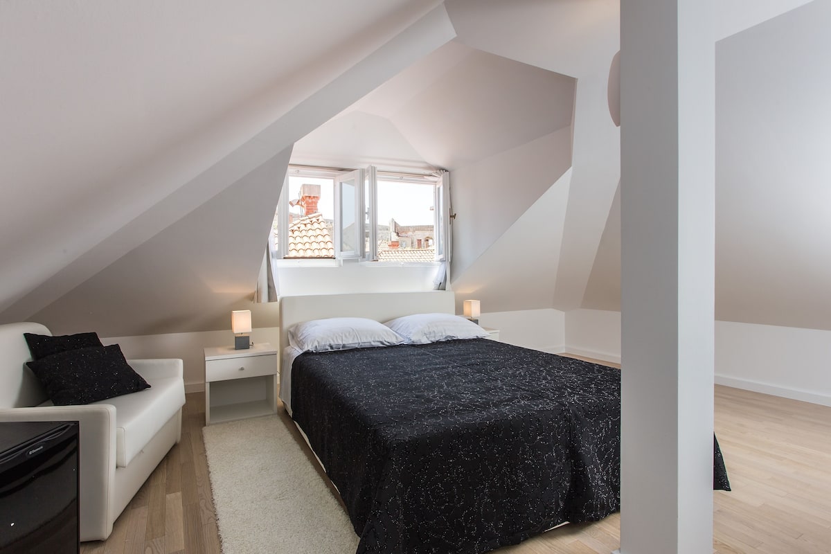 Um quarto com uma cama, mesa de cabeceira, cadeira e janela.