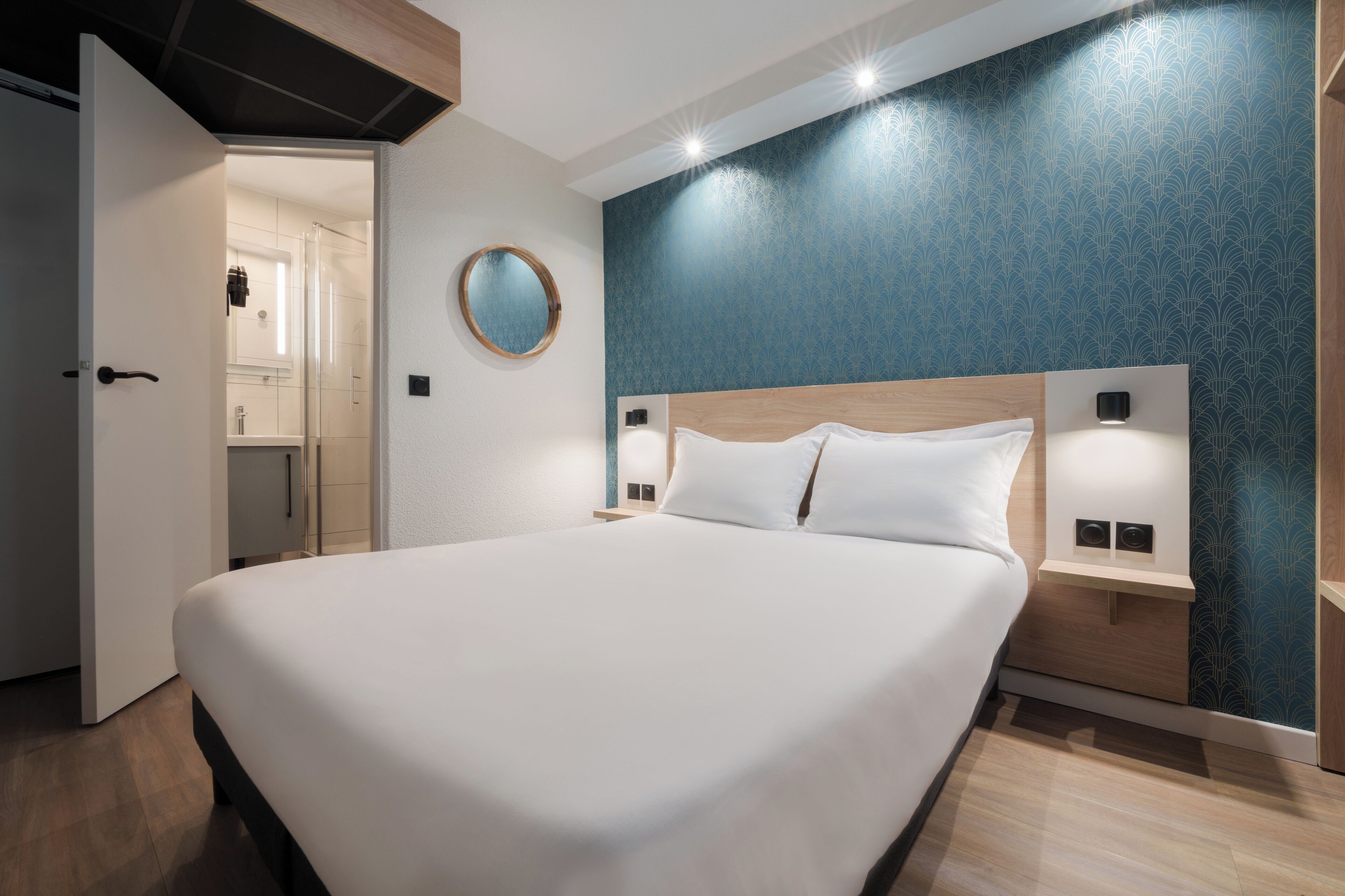 Quarto de hotel moderno com uma cama grande, cabeceira de madeira, espelho redondo e mesa de cabeceira embutida.