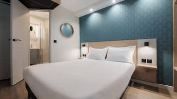 Quarto de hotel moderno com uma cama grande, cabeceira de madeira, espelho redondo e mesa de cabeceira embutida.