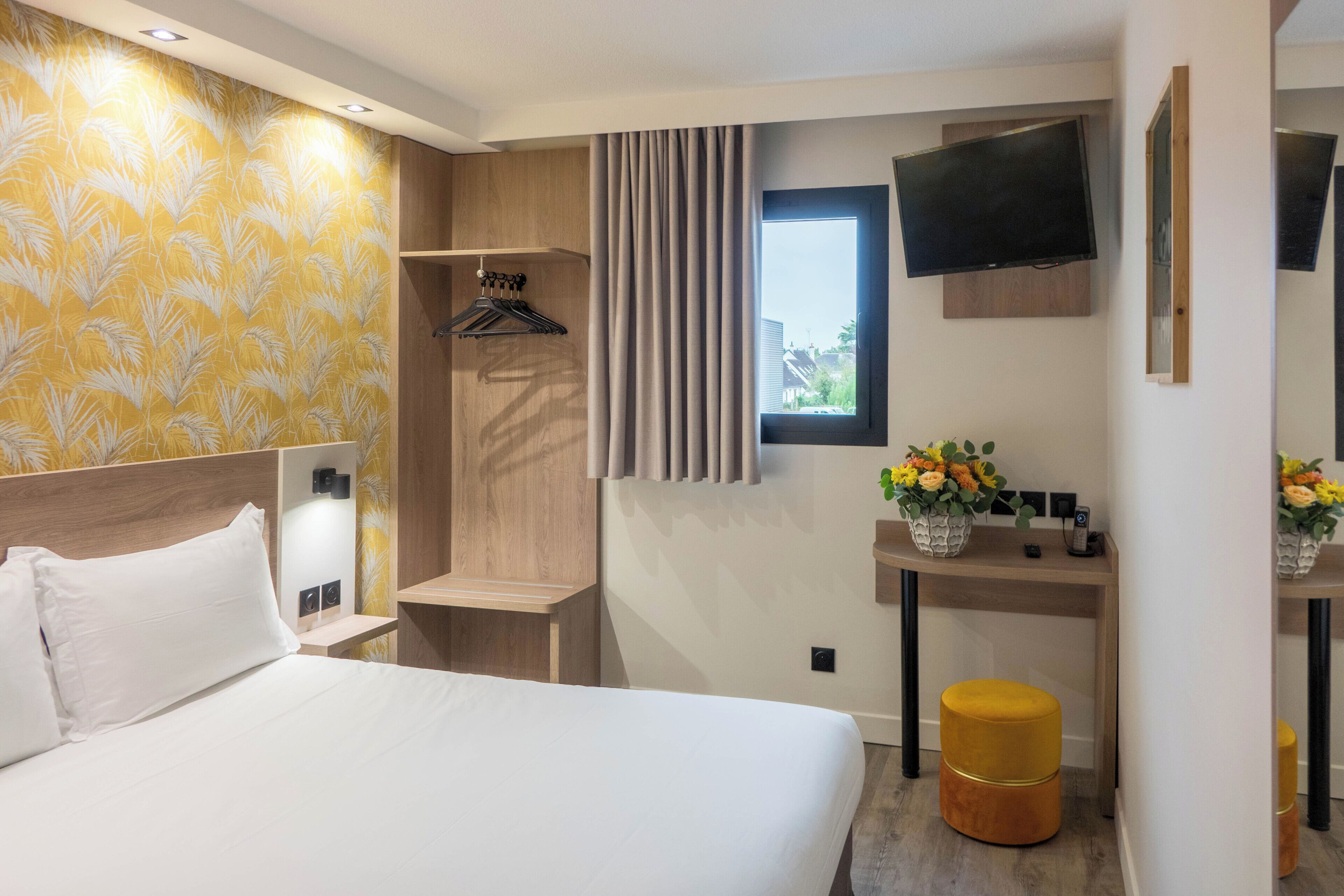 Quarto de hotel com uma cama, uma escrivaninha com televisão, uma janela e uma planta.
