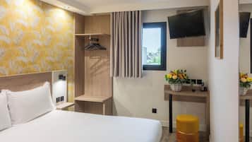 Quarto de hotel com uma cama, uma escrivaninha com televisão, uma janela e uma planta.