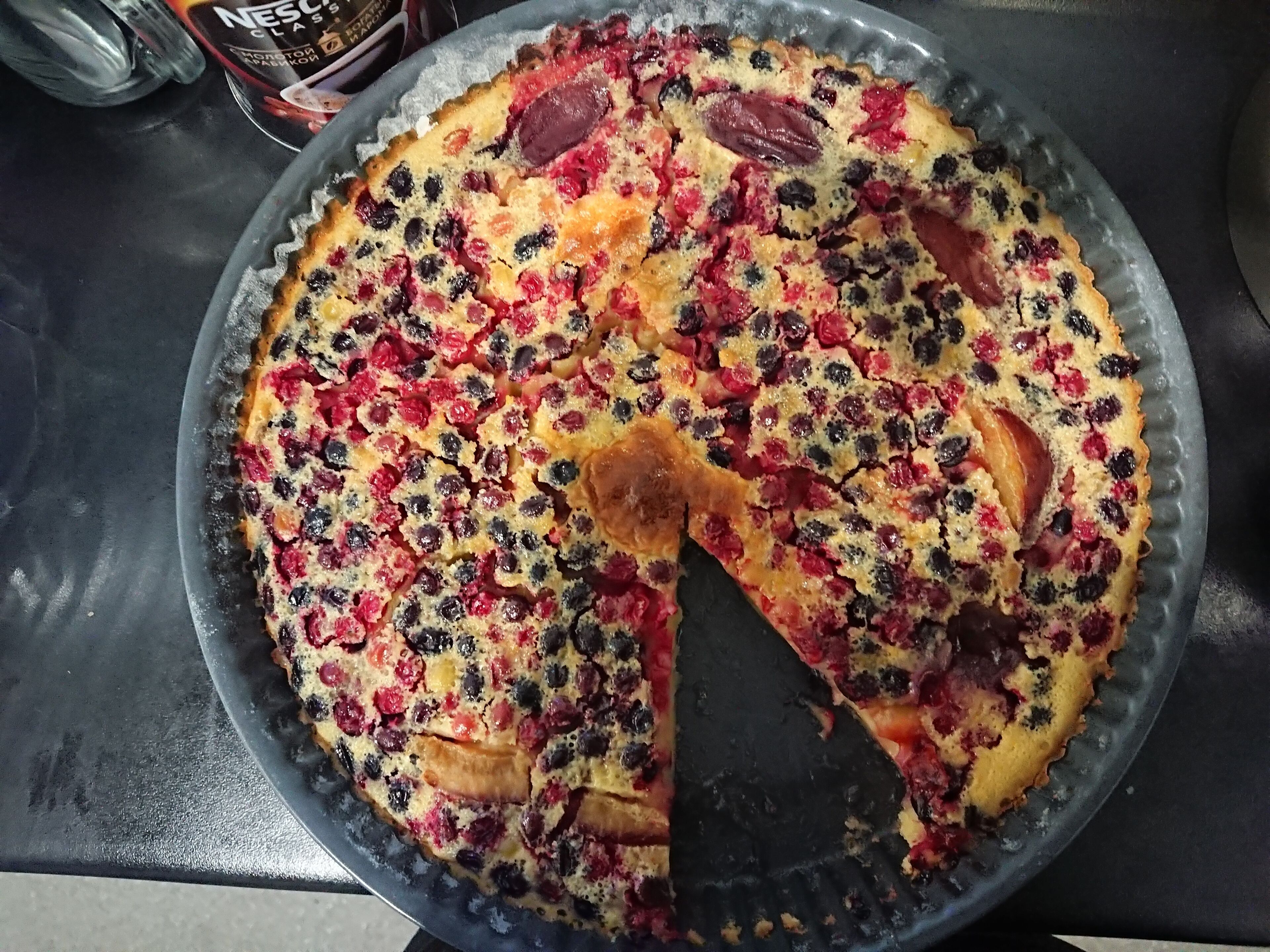 Uma travessa redonda de metal com uma fatia retirada, recheada com uma sobremesa coberta de frutas.