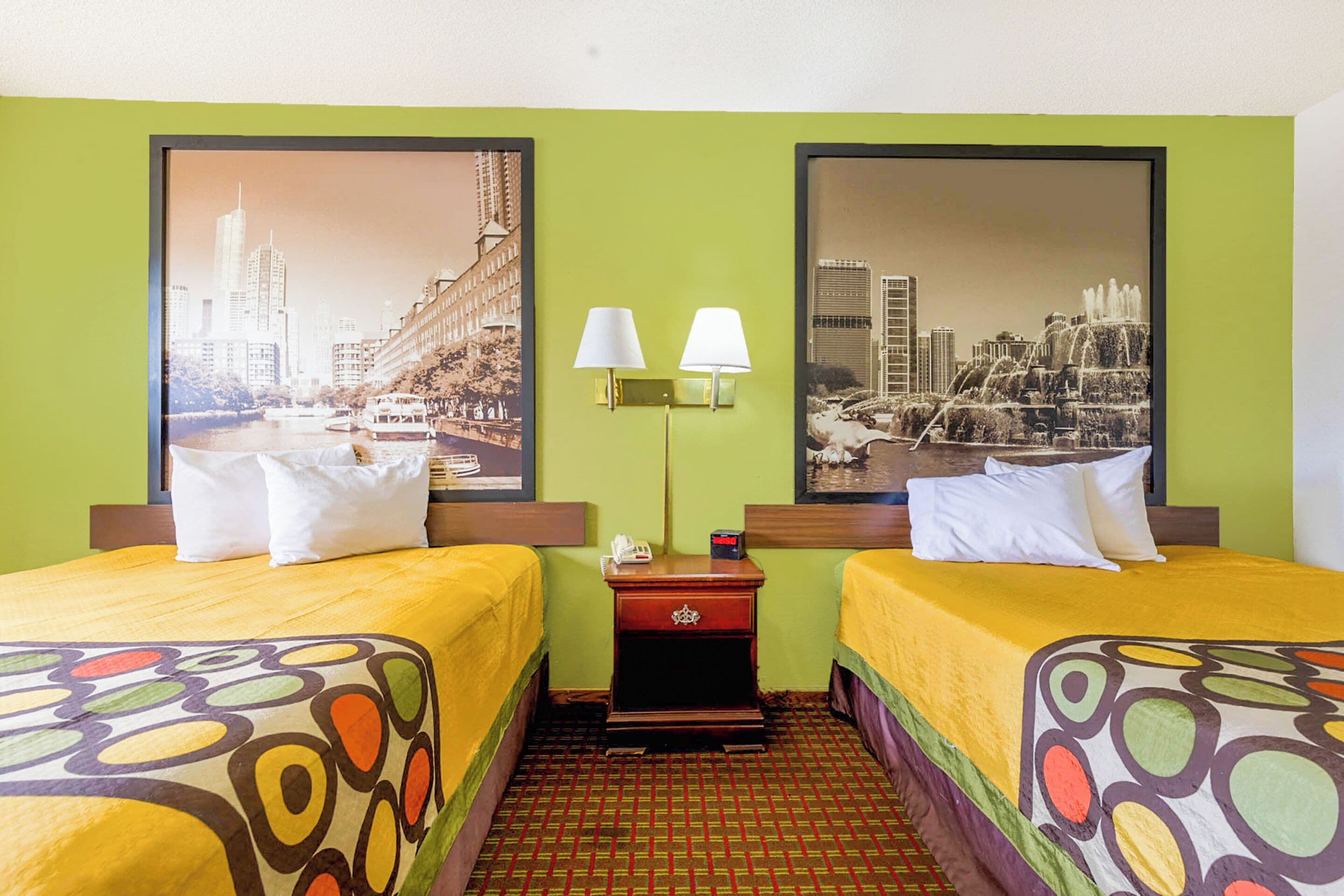 Quarto de hotel com duas camas, paredes verdes e cartazes com vistas da cidade.