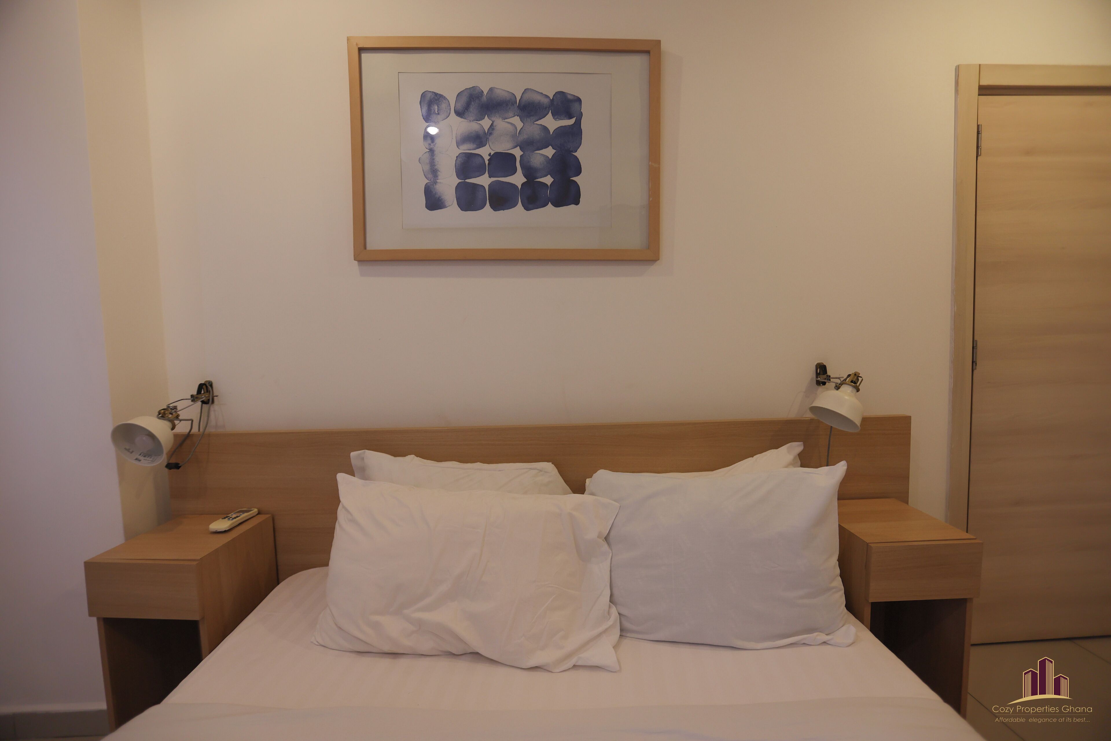 Cama bem arrumada com lençóis e almofadas brancas, uma obra de arte emoldurada acima e abajures em mesas de cabeceira de madeira.
