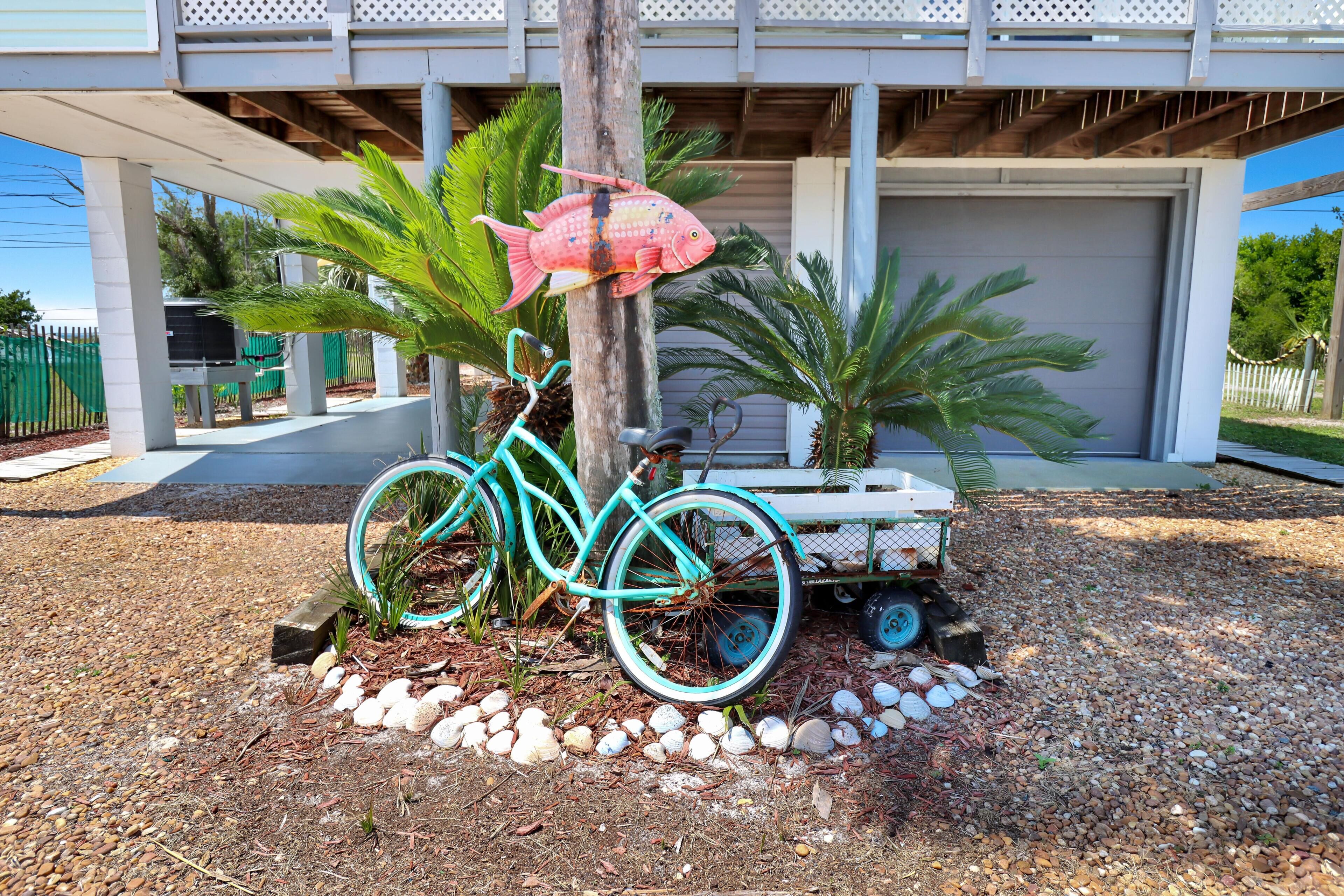 Uma bicicleta turquesa com decoração de peixe, uma palmeira e um carrinho branco com uma figura de peixe.