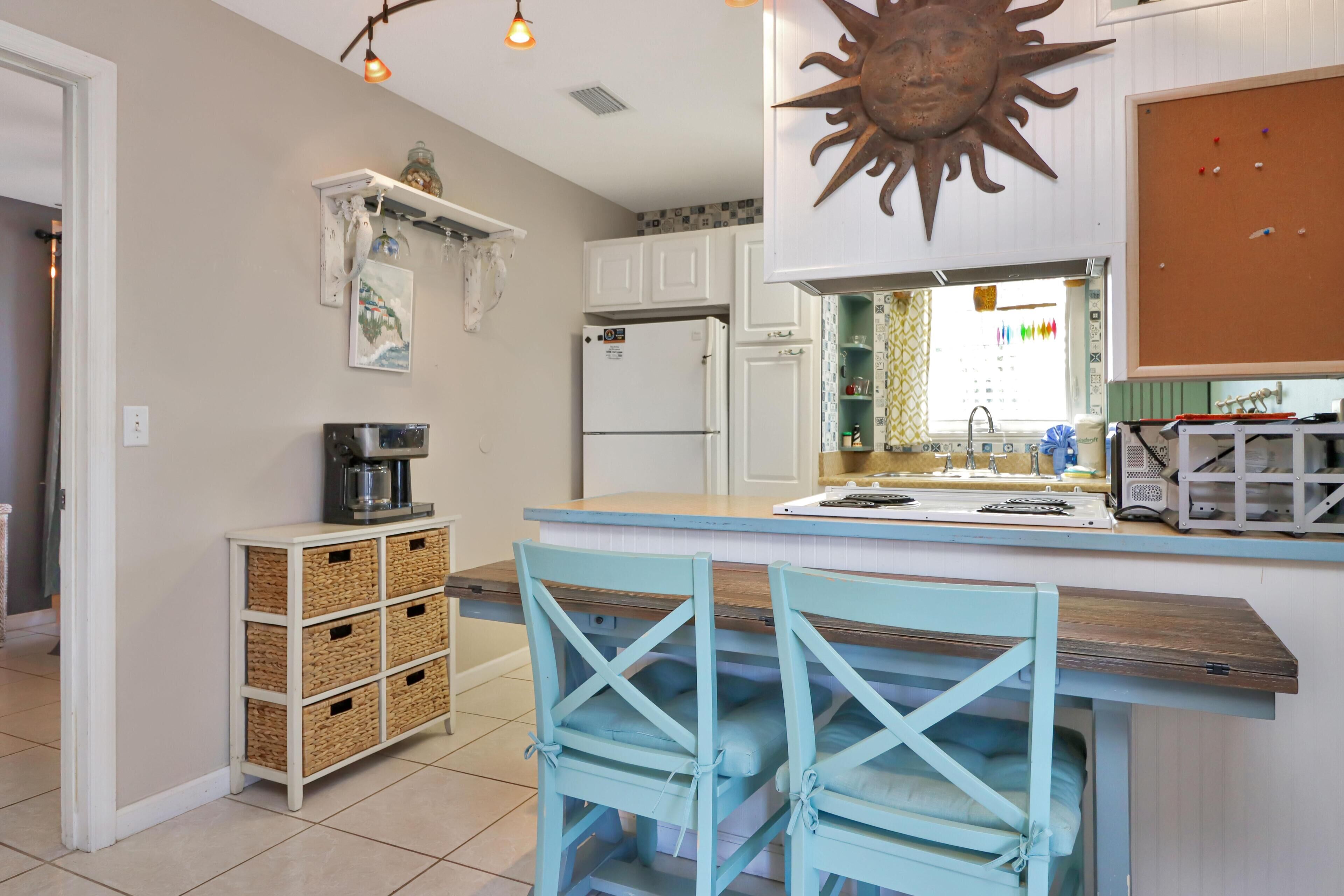 Uma cozinha com ilha central, armários brancos, uma cafeteira e uma decoração solar.
