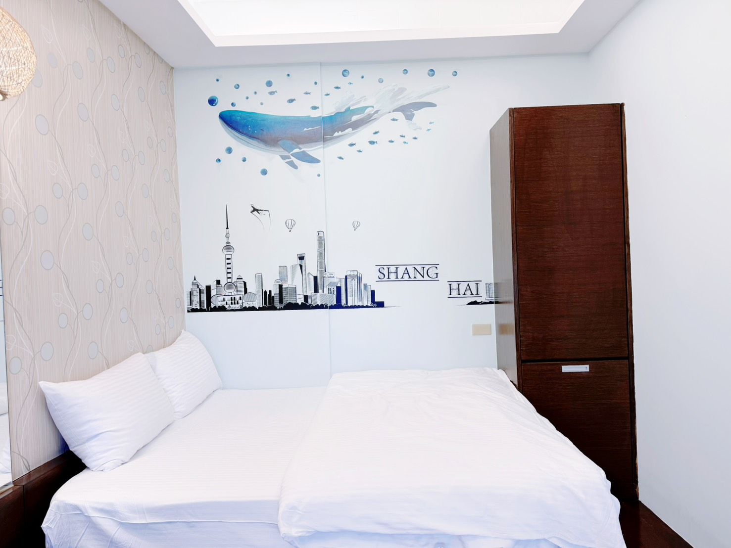 Quarto de hotel com uma cama, um guarda-roupa e um mural na parede retratando um horizonte urbano, com a inscrição 'SHANG HAI'.