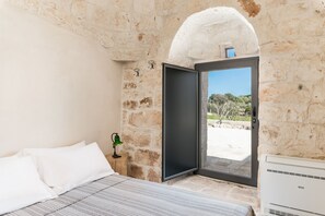 Quarto com parede de pedra, cama com roupa de cama branca, luminária verde e vista através de uma porta aberta.